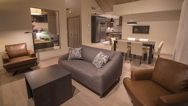 Uma sala de estar moderna com um sofá cinza, duas poltronas de couro marrom, uma mesa de centro escura e uma área de jantar com mesa e cadeiras.