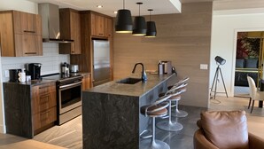 Uma cozinha moderna com armários de madeira, geladeira de aço inoxidável, forno embutido, cafeteira e um espaço bar com banquetas.