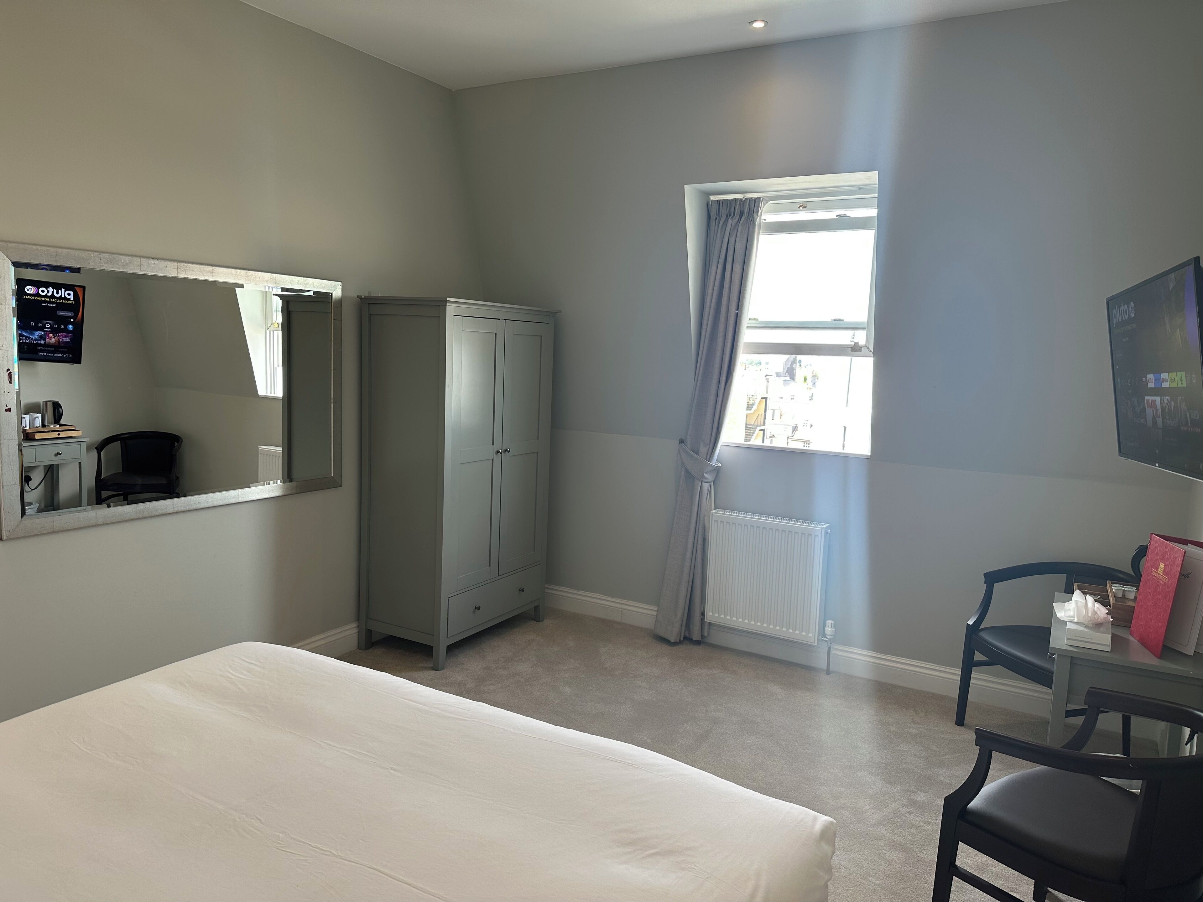 Quarto de hotel com cama, guarda-roupa, escrivaninha com cadeira, televisão e janela com cortinas.