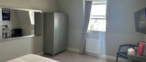 Quarto de hotel com cama, guarda-roupa, escrivaninha com cadeira, televisão e janela com cortinas.