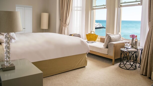 Quarto de hotel com uma cama grande, um sofá, uma mesinha com uma garrafa e um copo, e vista para o mar.