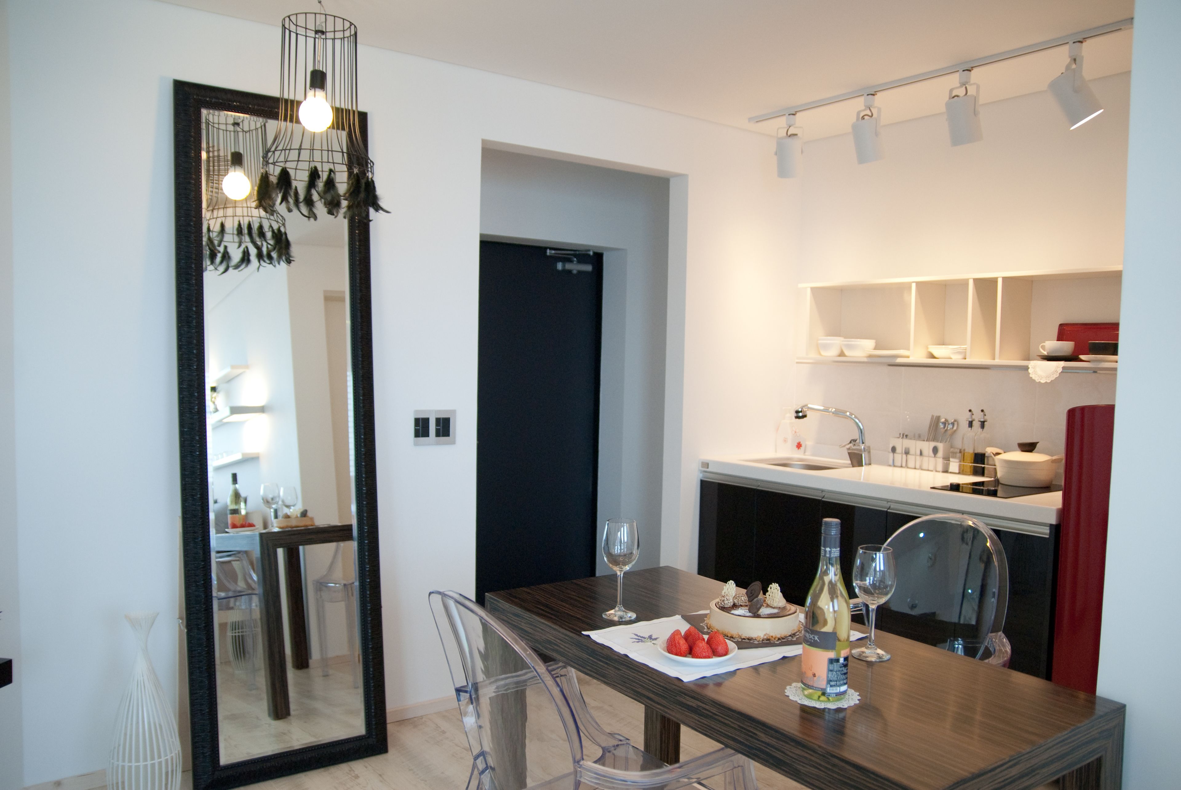 Uma sala de jantar moderna com mesa de madeira, cadeiras transparentes, um espelho grande e cozinha ao fundo.