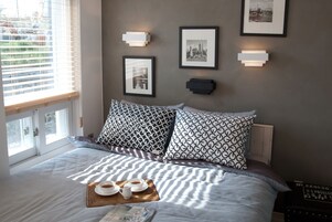 Uma cama bem arrumada com almofadas estampadas, um tabuleiro com xícaras e pires, e quadros nas paredes.