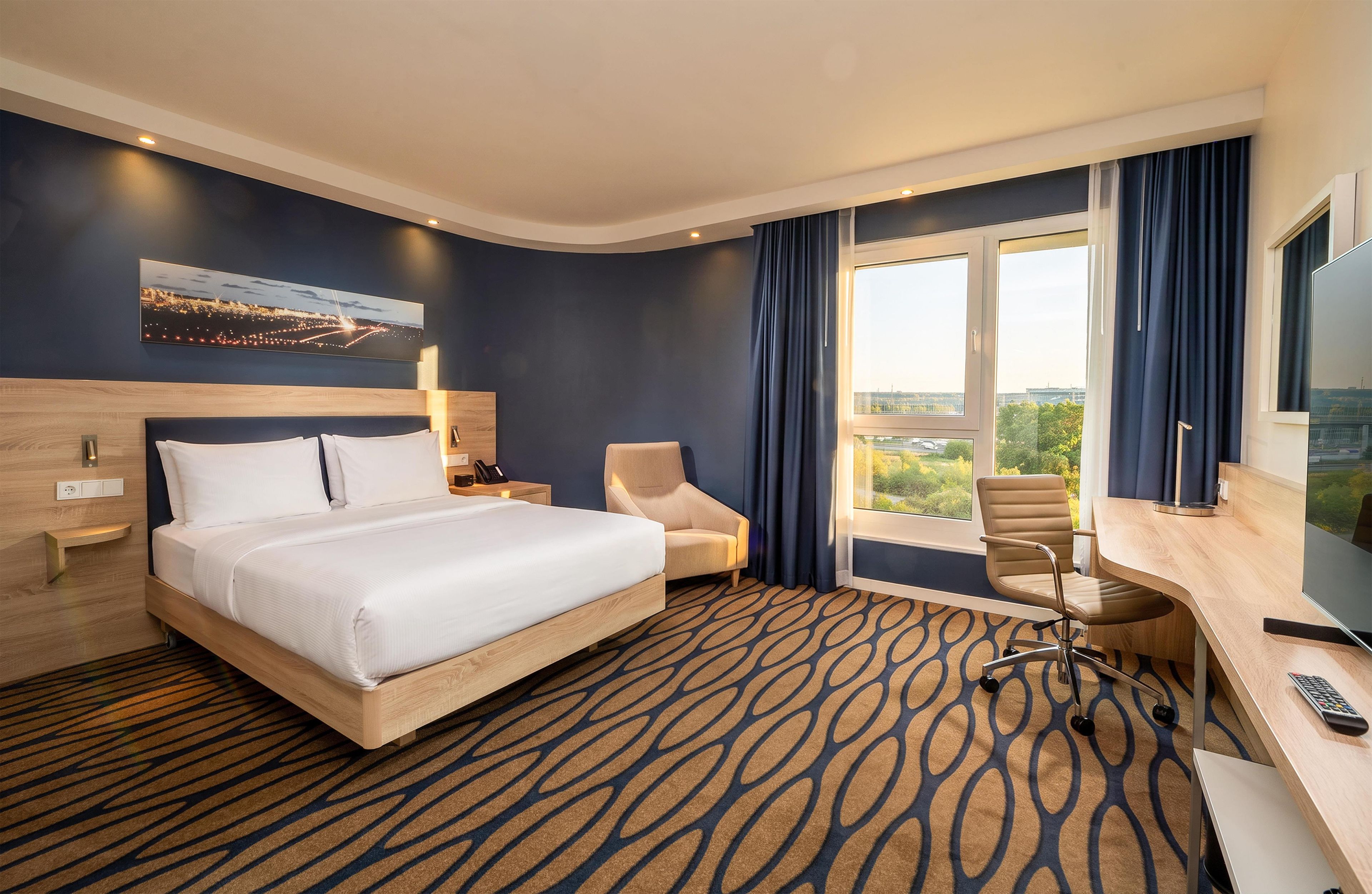 Quarto de hotel com uma cama grande, escrivaninha com cadeira, televisão e vista para a cidade pela janela.