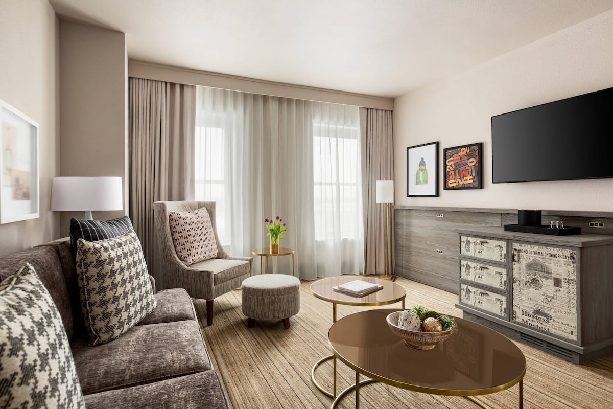 Quarto de hotel moderno com sofá, poltrona, banquinho, mesa de centro redonda e televisão de tela plana na parede.