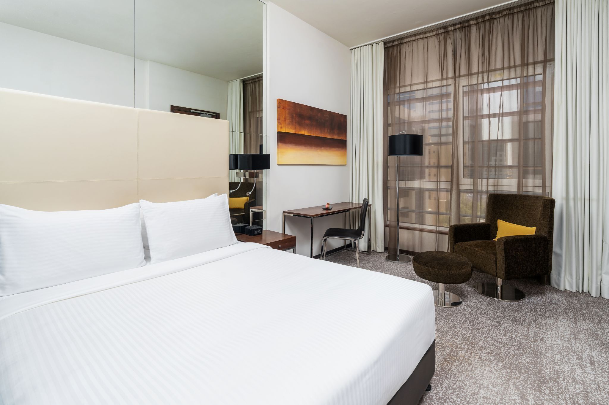Quarto de hotel com cama grande, escrivaninha, cadeira e vista da cidade através de cortinas translúcidas.
