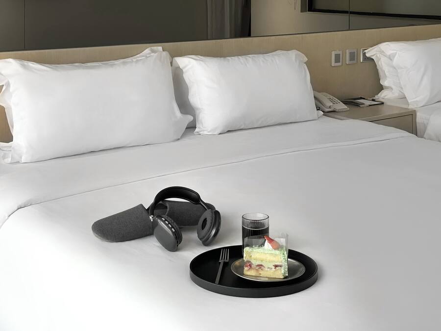 Cama bem arrumada com roupa de cama branca, um par de fones de ouvido, uma bandeja com sobremesa e um telefone ao lado da cama.