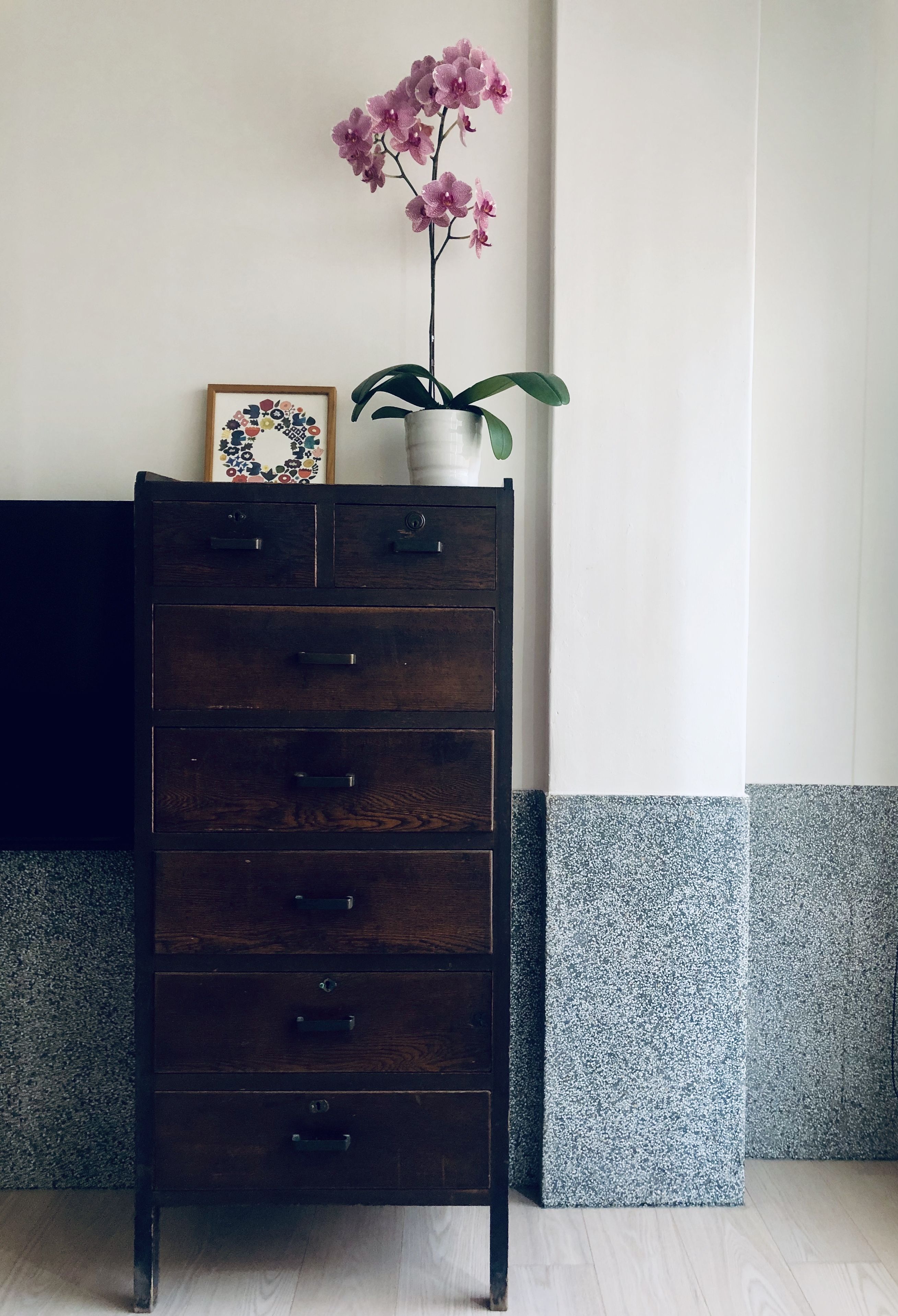 Uma cómoda de madeira escura com várias gavetas, um quadro emoldurado e uma orquídea num vaso, tudo em frente a uma parede com design de azulejos salpicados.