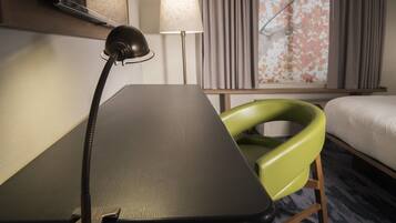 Quarto de hotel com uma escrivaninha, uma cadeira verde, uma cama, um abajur e uma janela com cortinas.