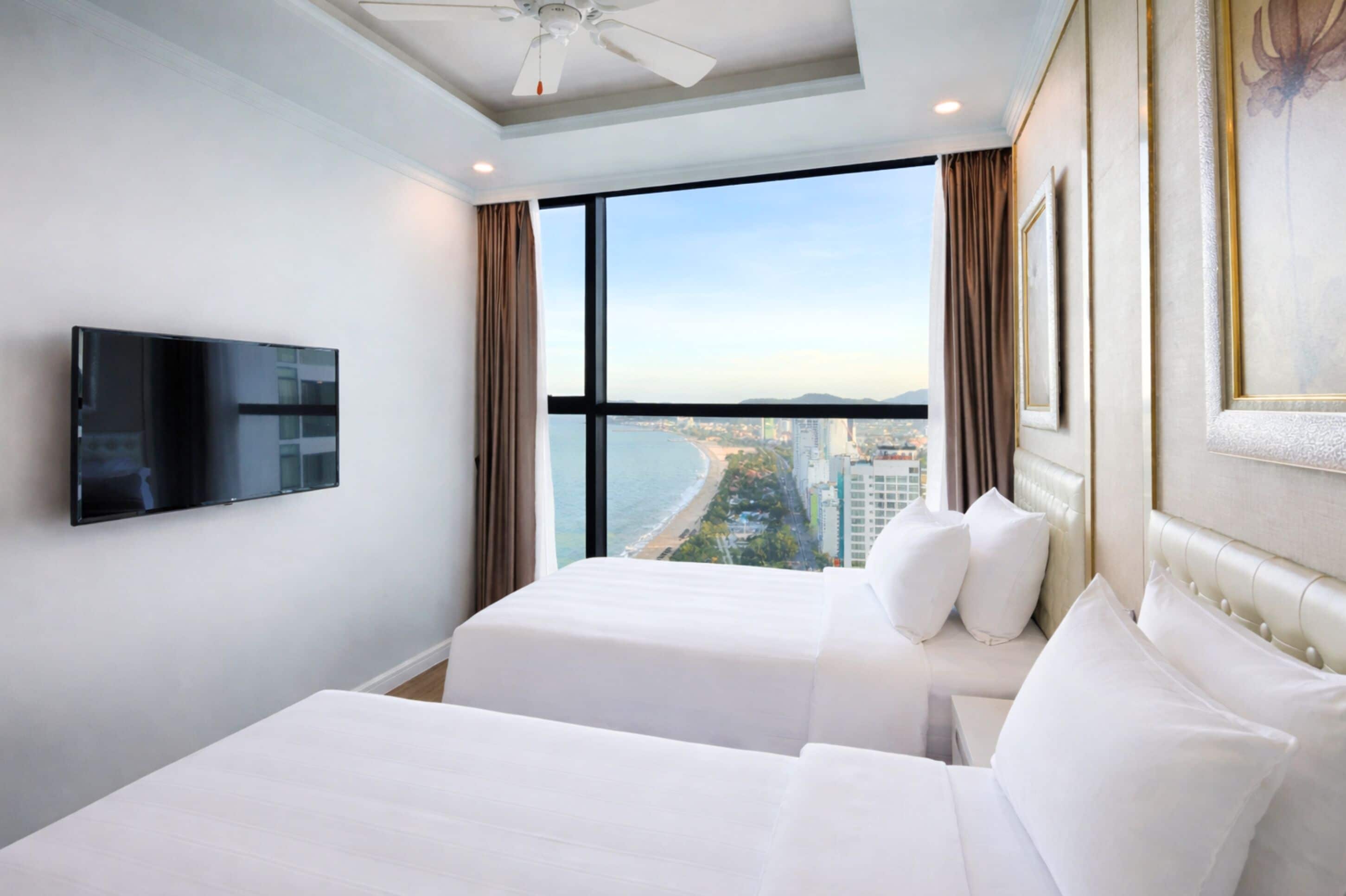 Quarto de hotel com duas camas, televisão, ventoinha no teto e uma grande janela com vista para a cidade.