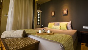 Cama bem arrumada com bandeja de xícaras brancas e um copo, um banco de fibra natural e uma janela grande com cortinas.