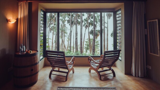 Quarto com duas cadeiras de madeira voltadas para uma janela ampla com vista para palmeiras altas.