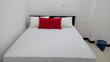 Uma cama de solteiro com roupa de cama branca e duas almofadas vermelhas, um cesto azul e uma parede branca.