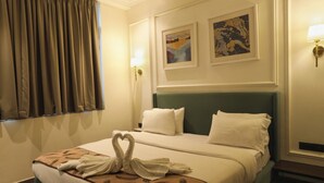 Quarto de hotel com cama, ventilador de teto, janela com cortinas e duas luminárias fixadas na parede.