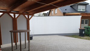 Uma área de varanda coberta com piso em padrão herringbone, uma mesa redonda e uma lixeira verde.