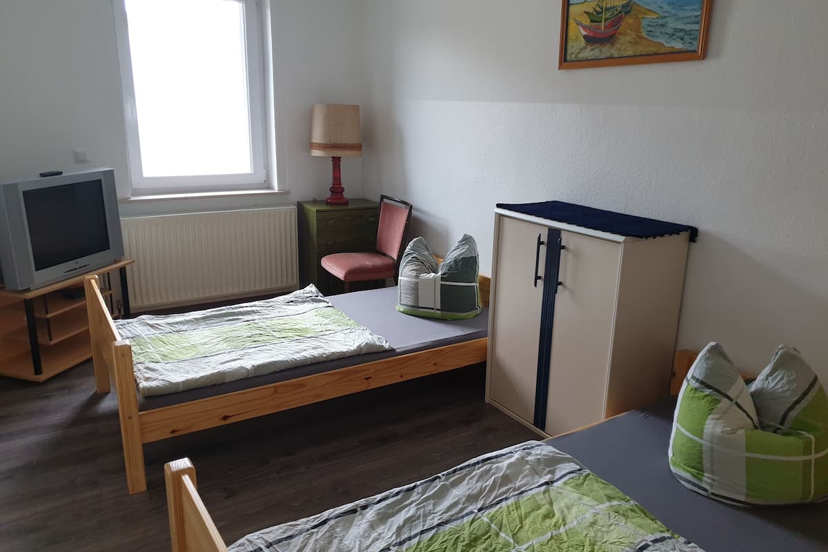 Um quarto pequeno com duas camas de solteiro, uma televisão, um armário, uma cadeira e uma janela.