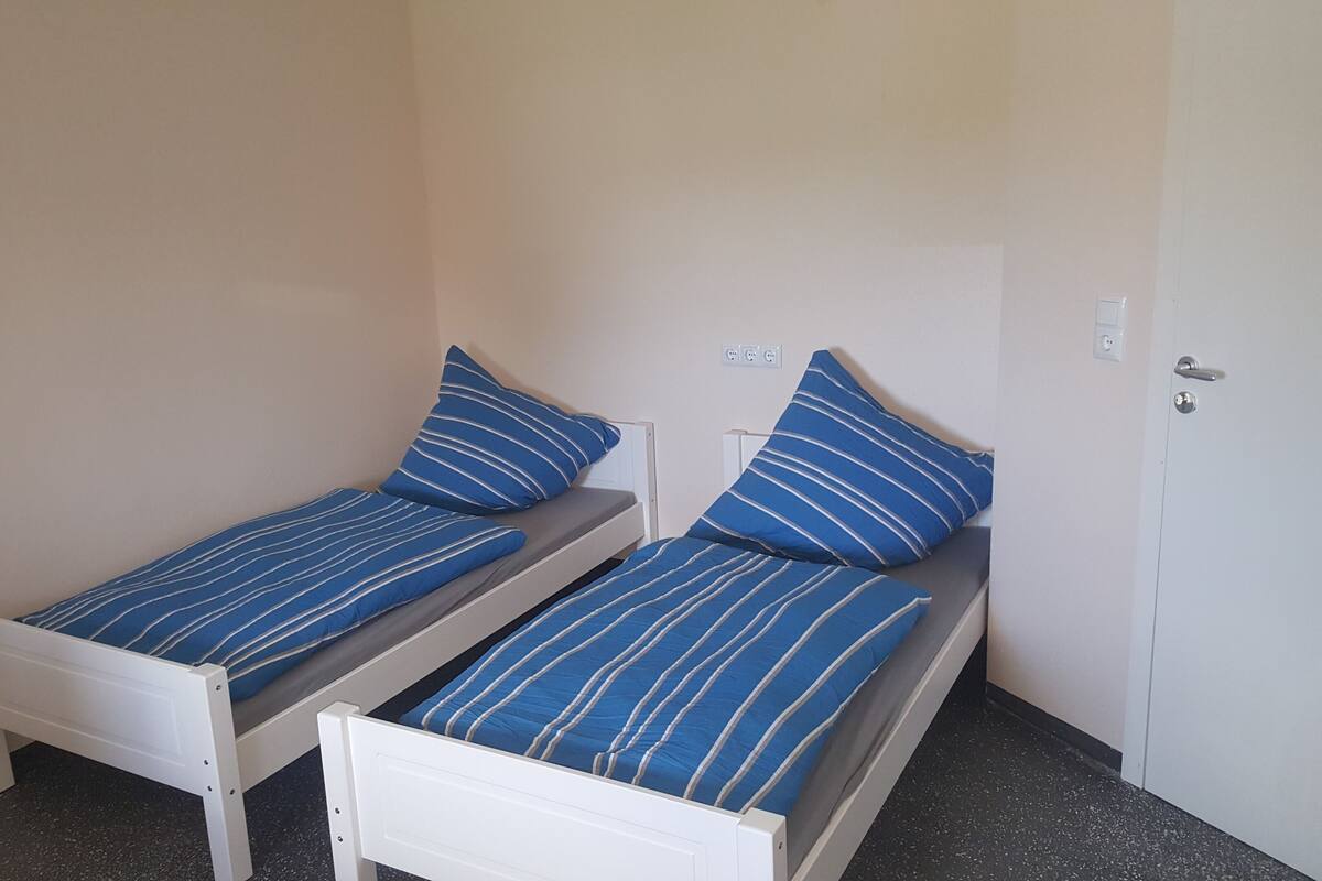 Duas camas de solteiro com roupa de cama listrada em azul num quarto pequeno com porta branca e parede de cor clara.