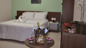 Quarto de hotel com cama, mesa de cabeceira, minibar e uma pequena mesa com uma garrafa de champanhe e duas taças.