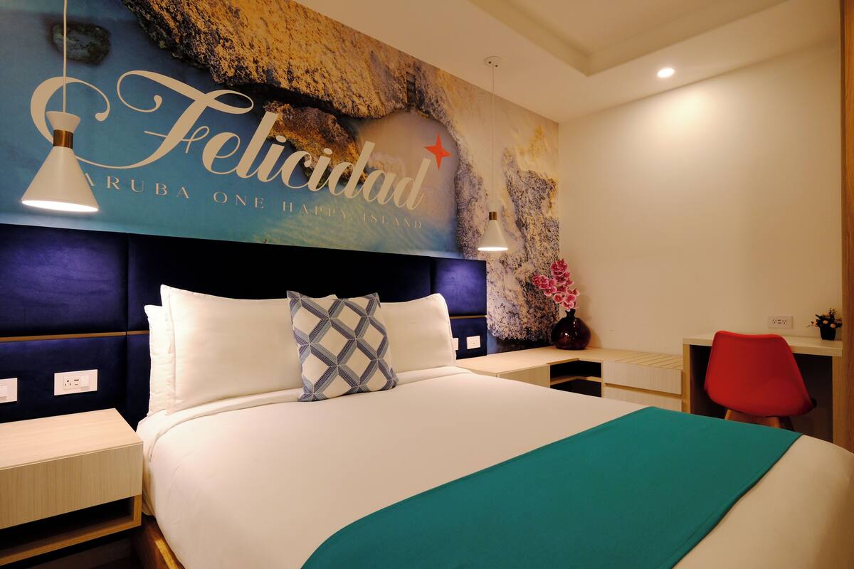 Quarto de hotel com uma cama, mesa de cabeceira, escrivaninha, cadeira e parede com um mural.
