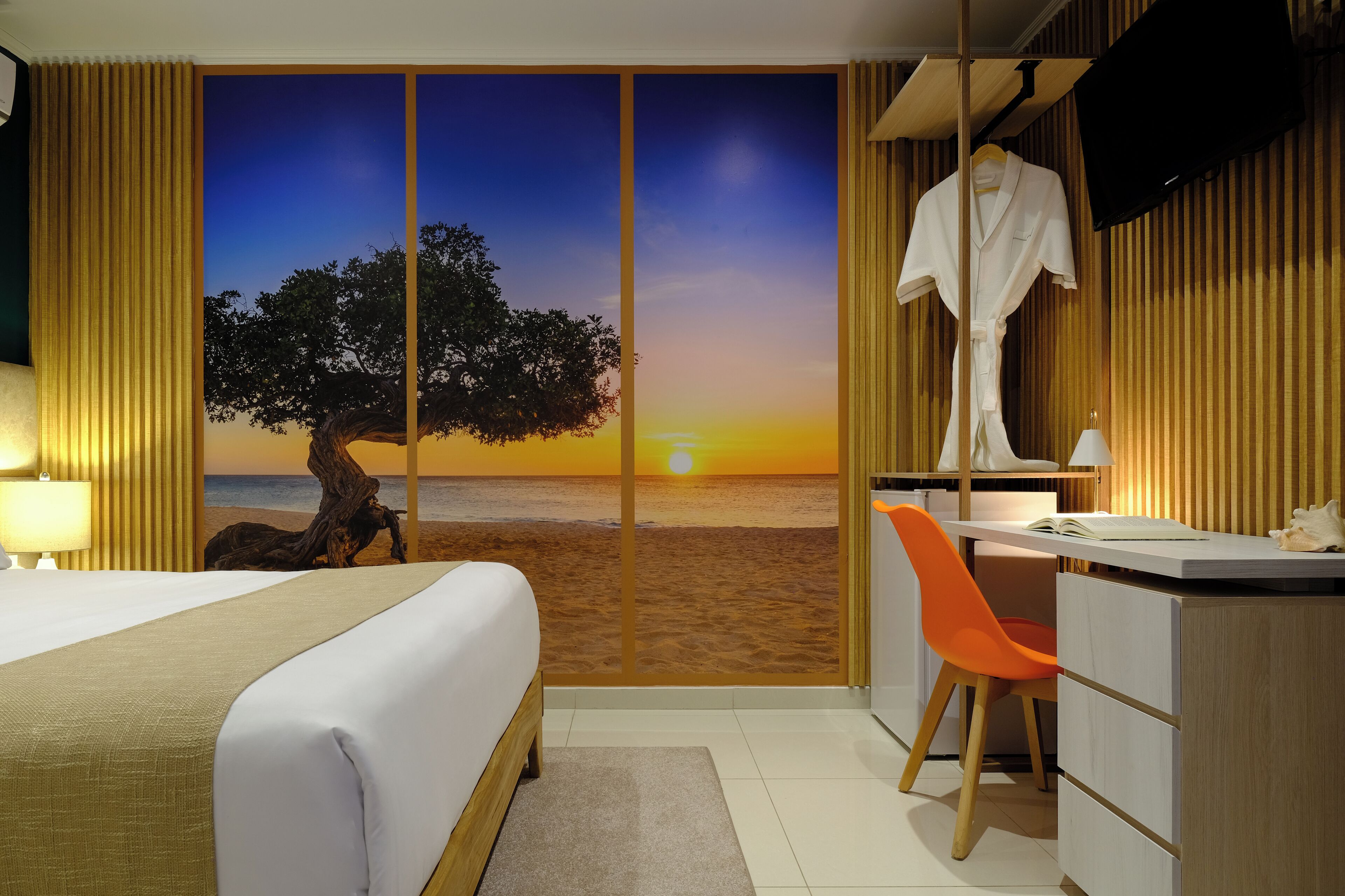 Quarto de hotel com cama, escrivaninha, cadeira e vista para a praia ao pôr do sol.
