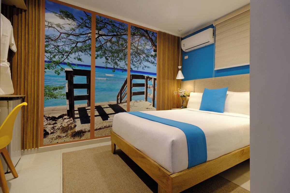 Quarto com cama, uma cadeira, escrivaninha e uma janela grande com mural de uma paisagem praiana.