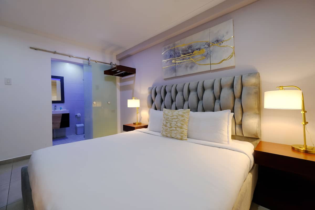 Quarto de hotel com uma cama grande, cabeceira, duas luminárias de apoio, um quadro abstrato na parede e um banheiro visível por uma porta aberta.