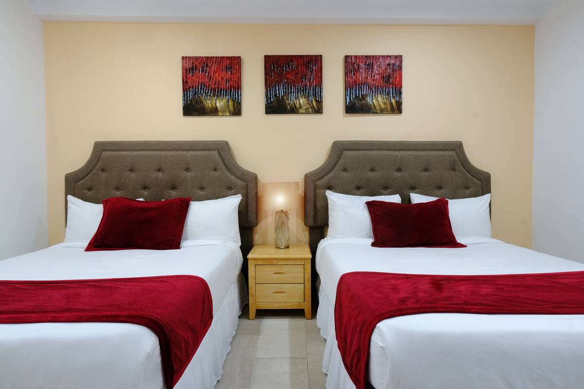 Quarto de hotel com duas camas, cada uma com manta vermelha e roupa de cama branca. Há três pinturas abstratas na parede, e uma mesa de cabeceira de madeira entre as camas.