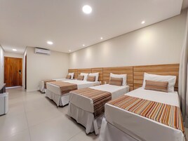 Quarto de hotel com quatro camas dispostas em linha, cabeceira de madeira e piso de azulejos brancos.