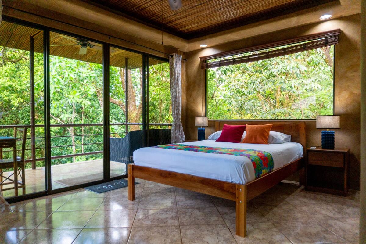 Quarto com cama grande, mesa de cabeceira e janela com vista para vegetação.
