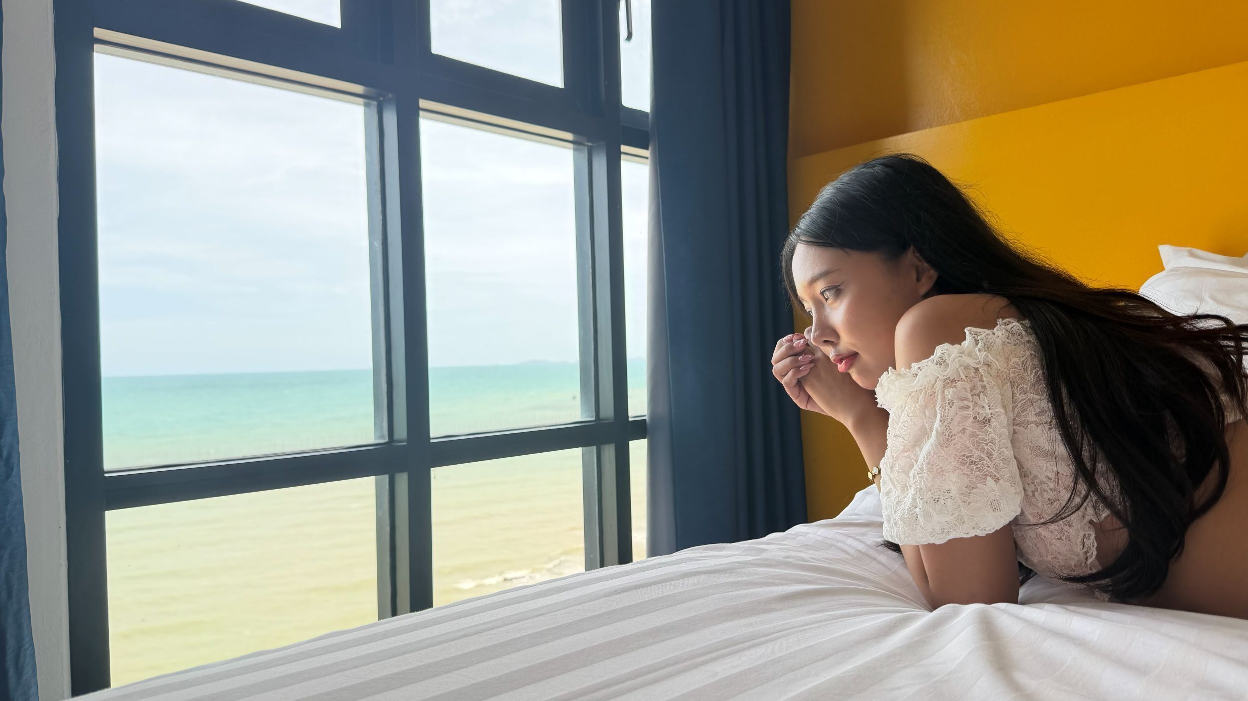 Quarto com uma grande janela com vista para uma praia, uma cama com roupa de cama branca e uma parede amarela.