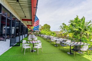 Área externa com mesas e cadeiras, cercada por vegetação e um edifício com grandes janelas.
