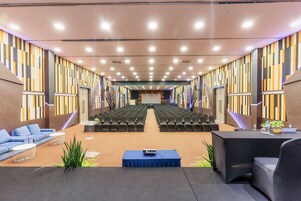 Sala de conferências com fileiras de cadeiras, palco e área de estar com sofás e mesas.