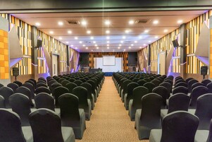 Sala de conferências com fileiras de cadeiras, palco com tela e sistema de som.
