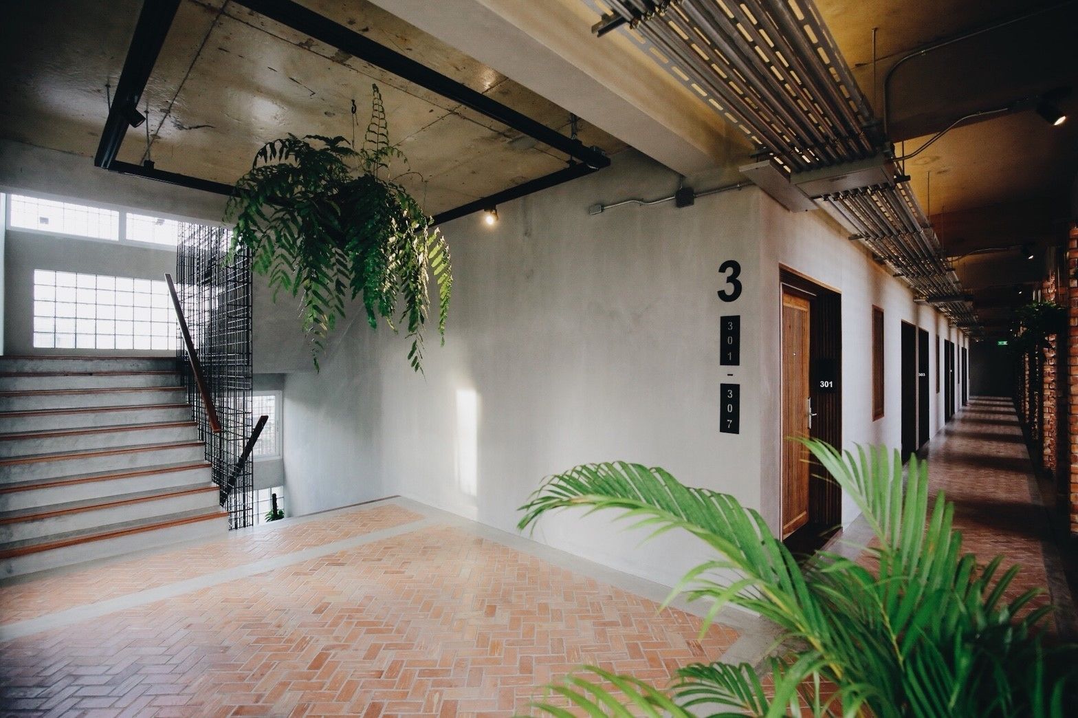 Um corredor amplo com piso de tijolos, escada com grades de metal e plantas penduradas.