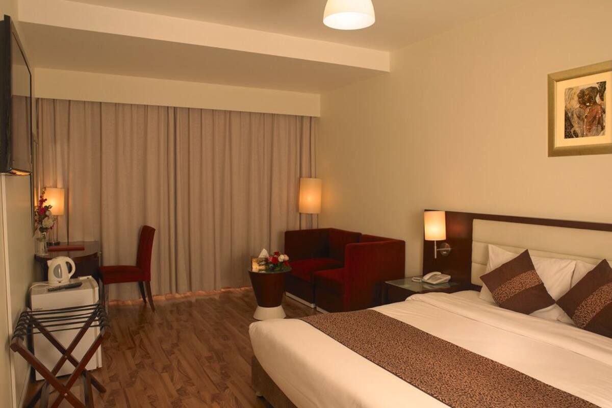 Quarto de hotel com cama, escrivaninha com cadeira, sofá, luminária e um quadro na parede.