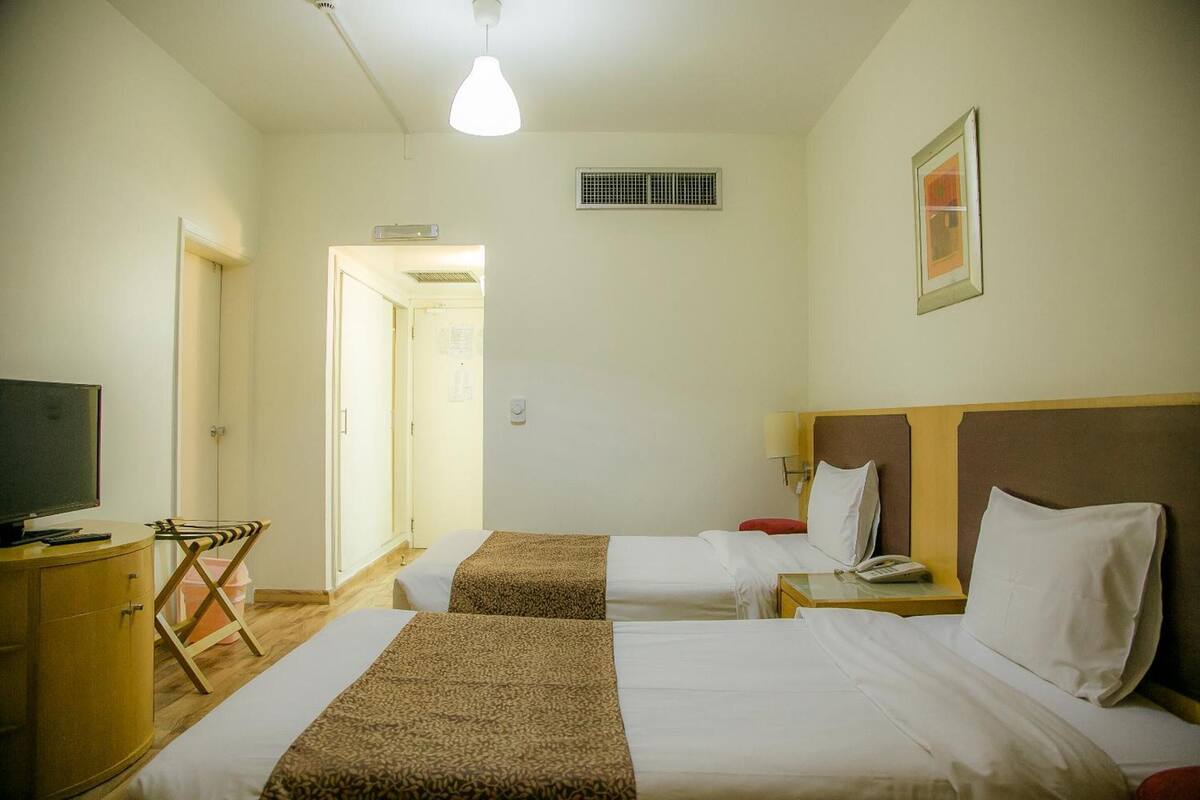 Quarto de hotel com duas camas, uma escrivaninha com candeeiro, televisão e janela com persianas.