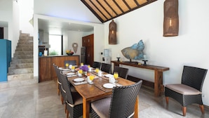 Uma sala de jantar com mesa de madeira posta para seis pessoas, cercada por cadeiras de vime, e uma área de cozinha com um dispensador de água e uma tigela de frutas à vista.