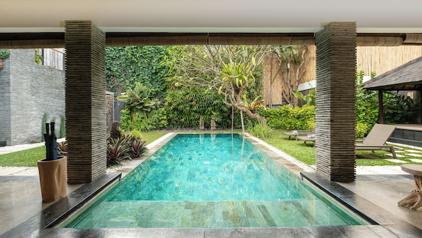 Uma piscina retangular com água azul cristalina, rodeada por um deck de pedra e ladeada por um sebe baixo.