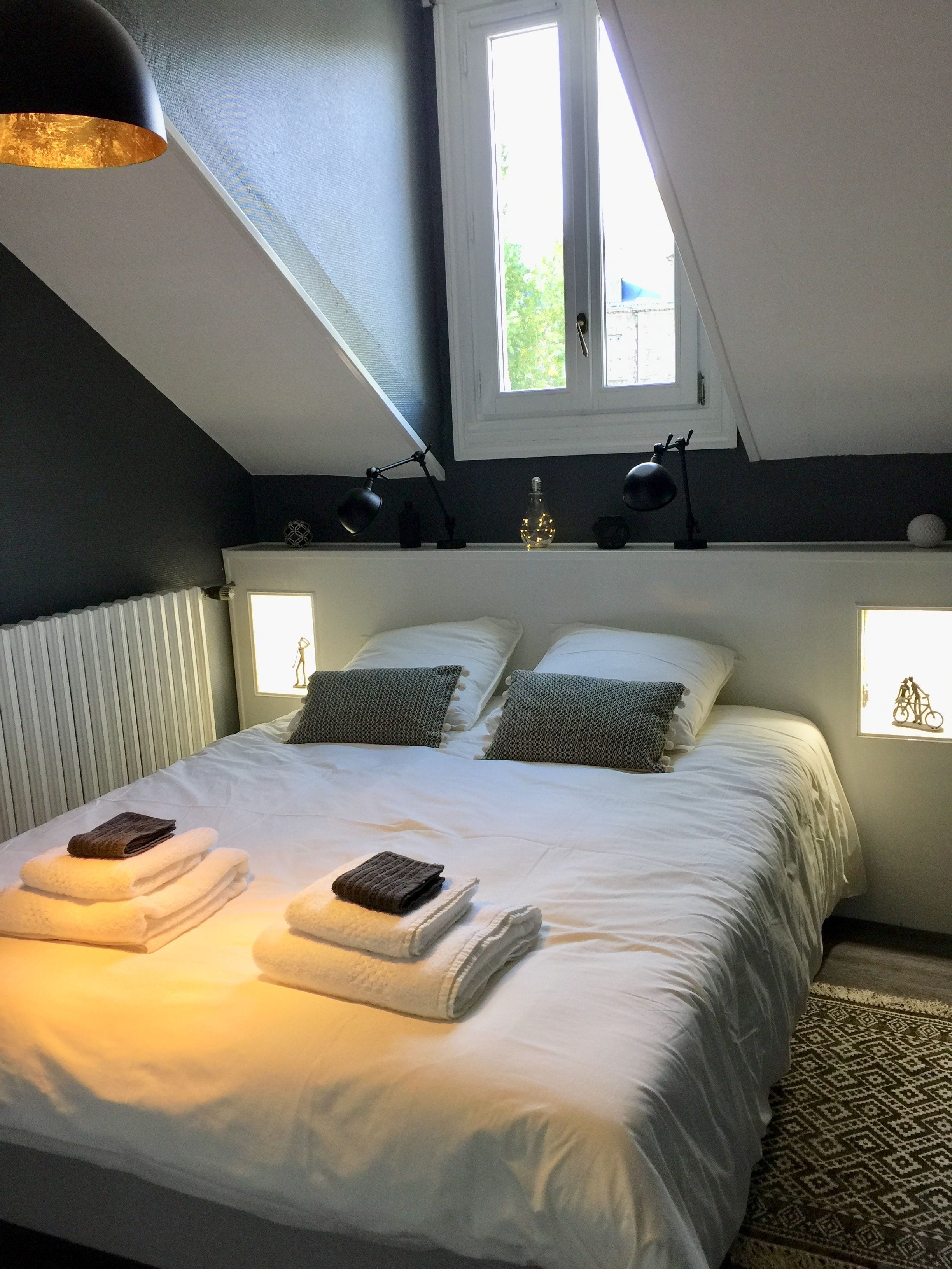 Quarto com teto inclinado, cama com roupa de cama branca, duas luminárias de cabeceira e janela com vista para o verde.