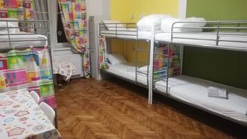 Quarto tipo dormitório com beliches, piso de madeira em padrão herringbone e uma mesa com toalha de mesa florida.