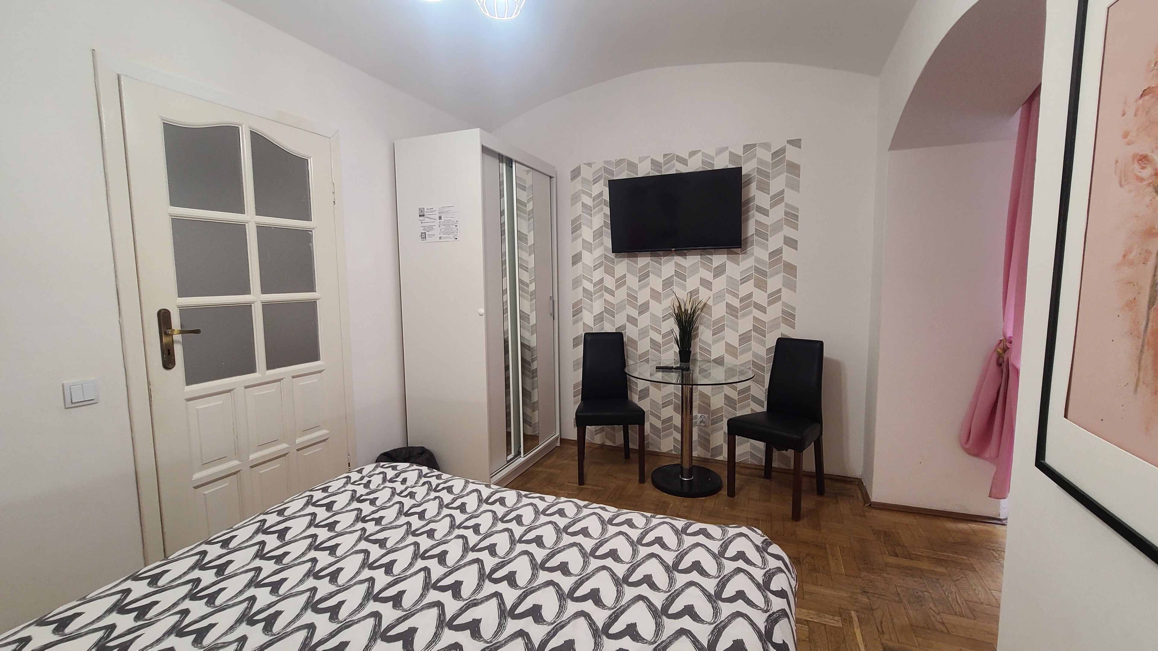 Quarto com uma cama, televisão presa na parede, mesa pequena com duas cadeiras e porta com painéis de vidro jateado.