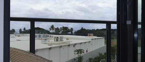 Varanda com cadeira de vime e uma mesa pequena, com vista para um terraço com edifícios e palmeiras.