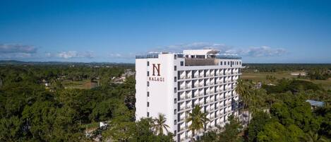 Um hotel moderno e alto, com um logotipo 'N' bem visível na fachada, cercado por vegetação exuberante e um céu azul límpido.