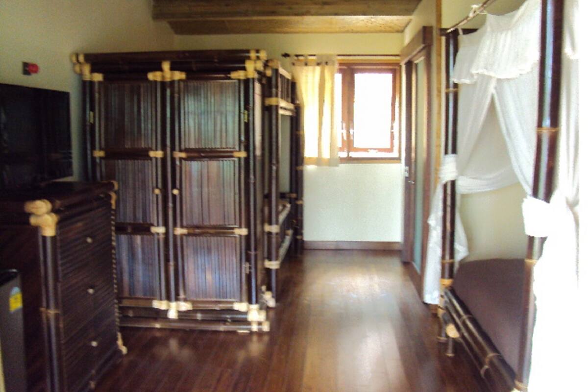 Quarto com piso de madeira, cama com dossel, guarda-roupa e televisão sobre um suporte.