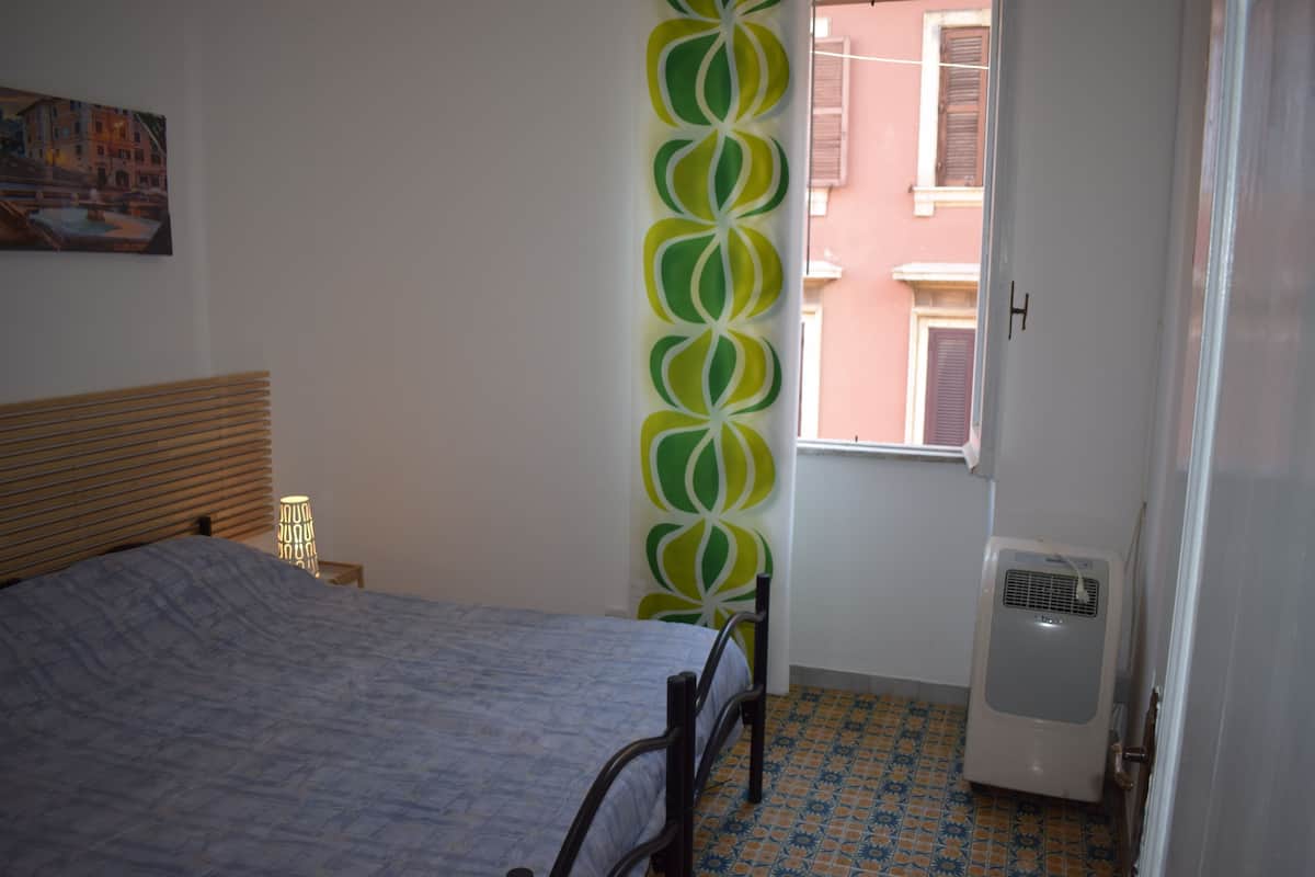 Quarto com cama, radiador e janela com vista para edifícios.
