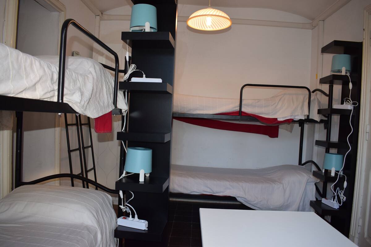 Quarto com beliches, uma escrivaninha e estantes, cada um com luminária e tomadas.