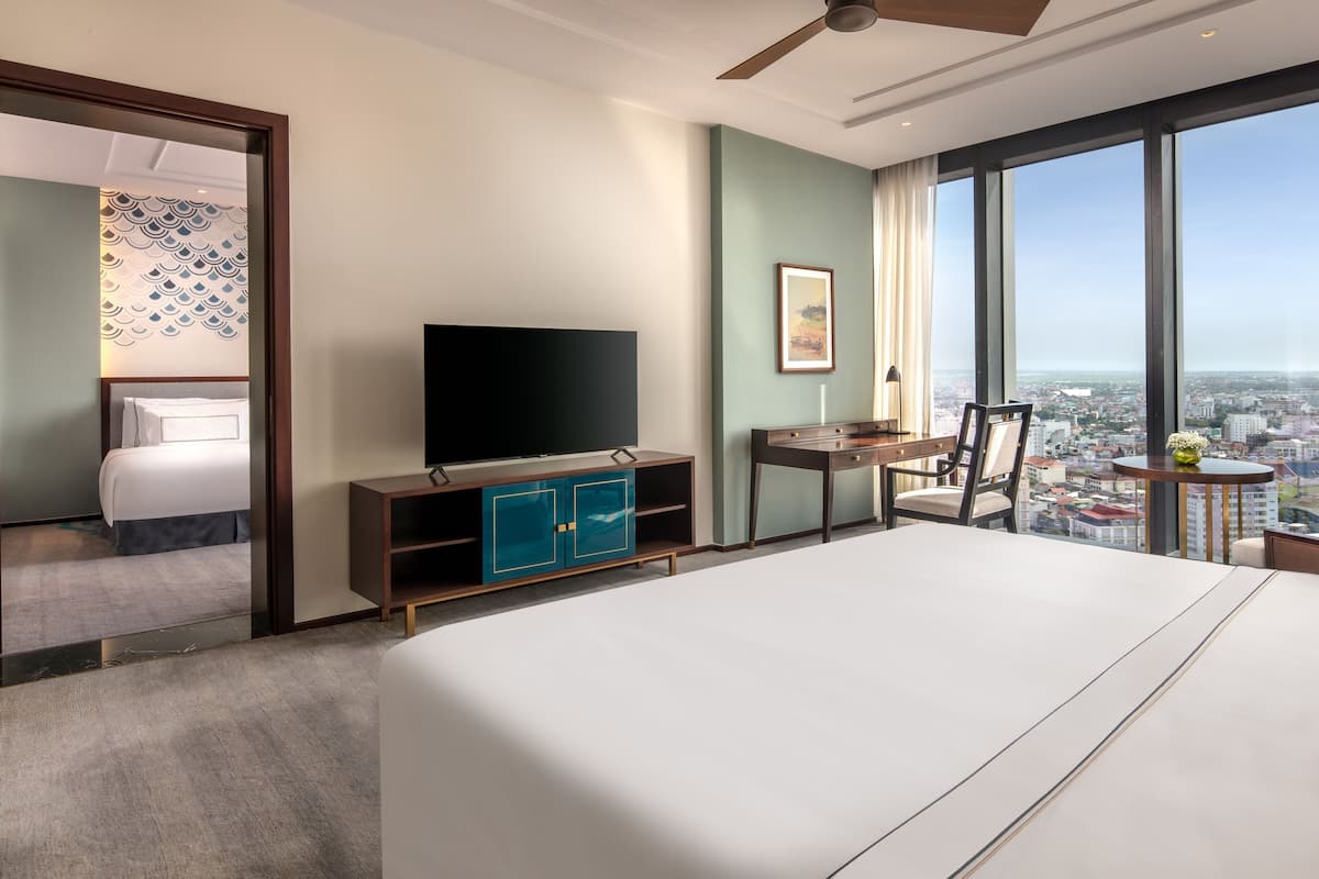 Quarto de hotel com uma cama grande, uma TV em um móvel de madeira, uma escrivaninha com cadeira e vista para a cidade através de amplas janelas.