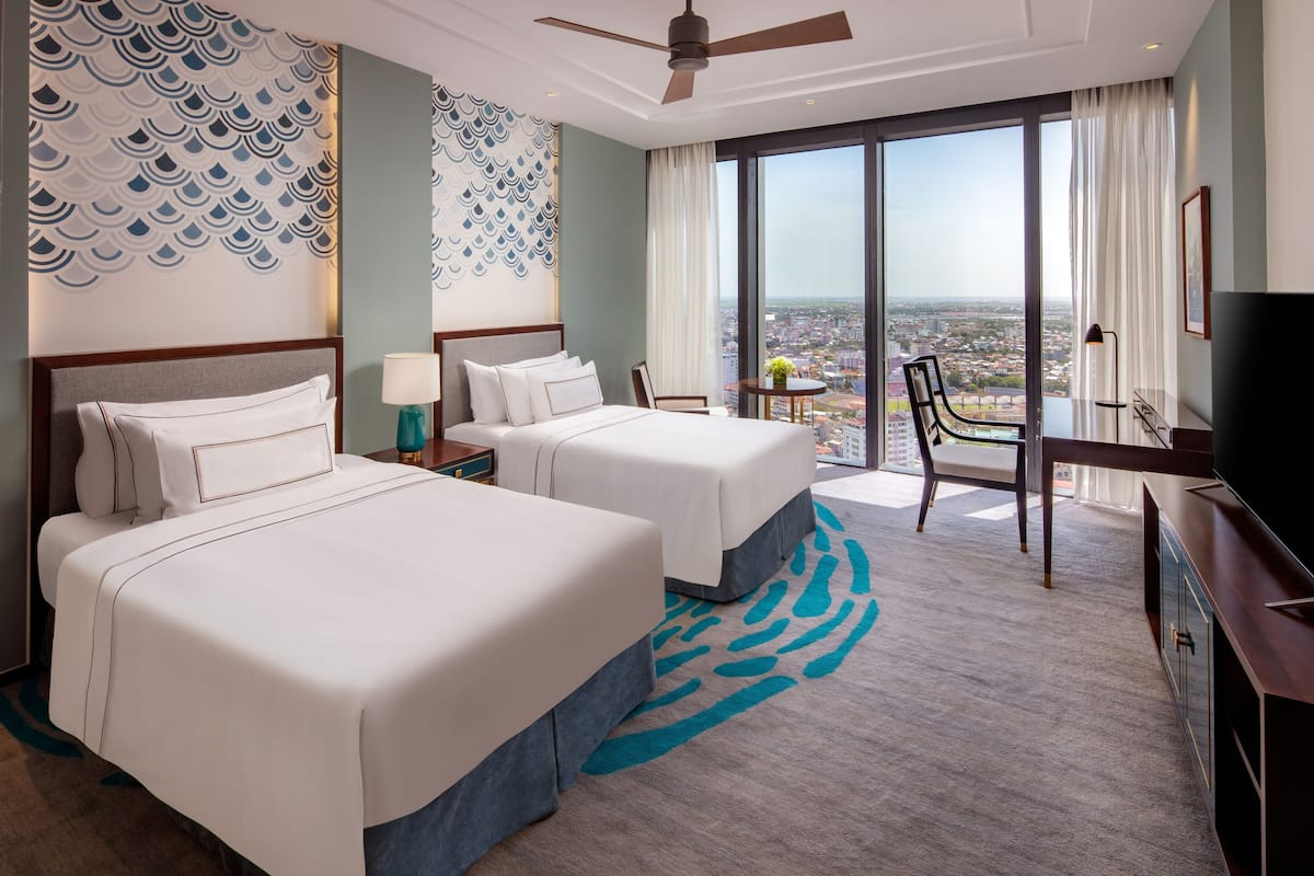 Quarto de hotel com duas camas, uma escrivaninha, uma cadeira, uma TV e uma janela grande com vista para a cidade.