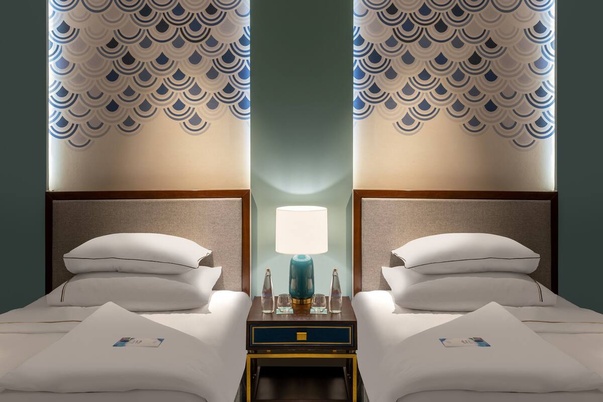 Quarto de hotel com parede espelhada, cama com roupa de cama branca, mesa de cabeceira com abajur e apoio com duas taças.