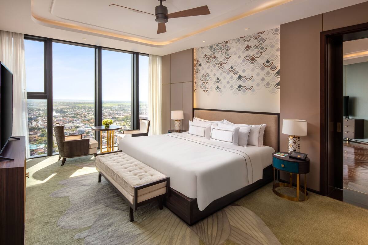 Quarto de hotel moderno com uma cama grande, um espaço de estar com um banco, uma mesinha e uma janela ampla com vista para a cidade.